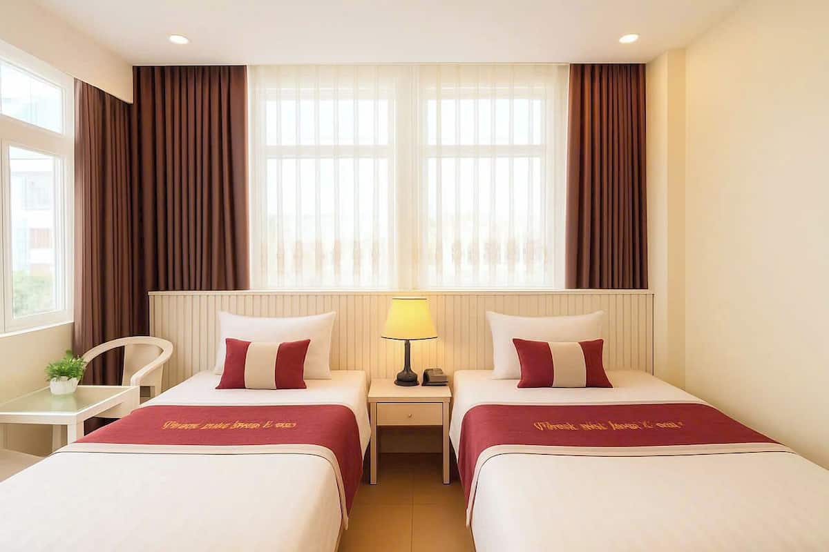 Quarto de hotel com duas camas, uma mesinha de cabeceira com abajur, uma pequena mesa com uma planta e uma janela com cortinas.