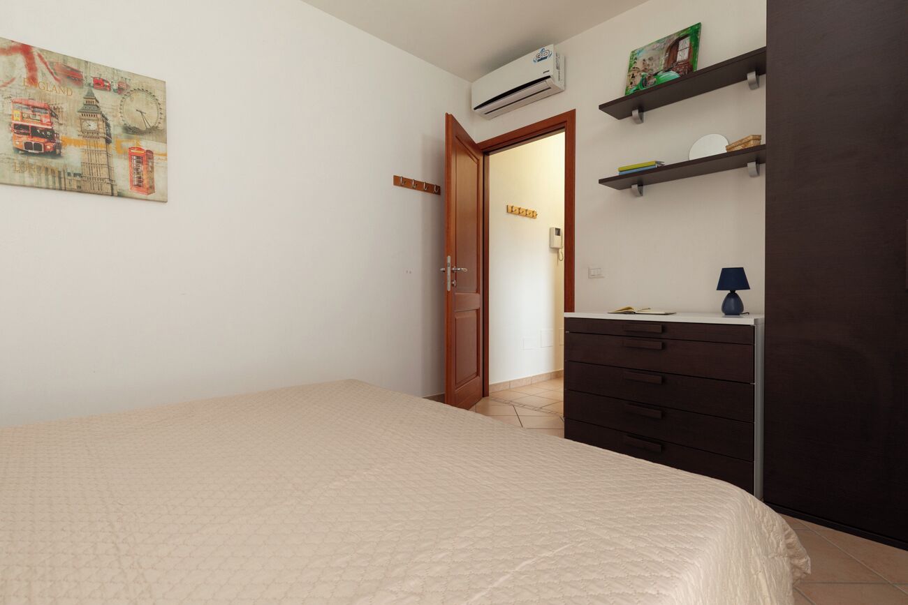 Quarto com cama, cômoda, escrivaninha com luminária, pintura de pontos turísticos de Londres e aparelho de ar condicionado.