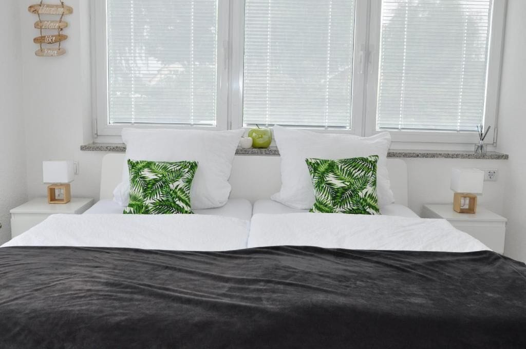 Cama bem arrumada com roupa de cama branca e almofadas com padrão de folhas verdes, mesa de cabeceira com abajur e janela com persianas.
