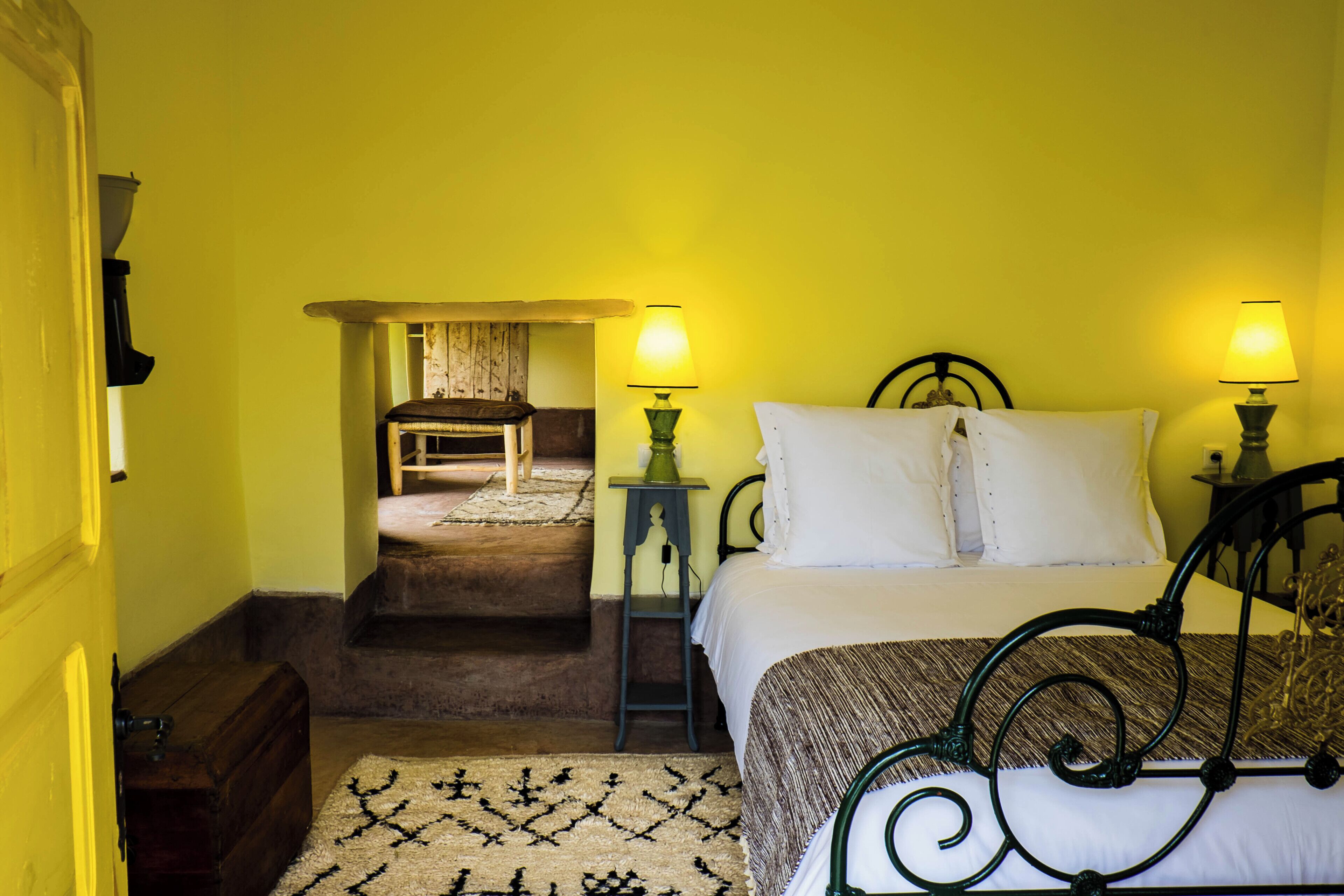 Quarto com cama, duas luminárias de cabeceira, um baú de madeira, um tapete estampado e uma escada que leva a uma pequena mesa com uma cadeira.