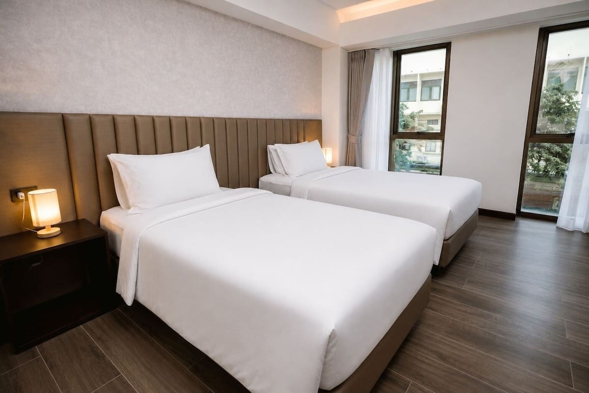 Quarto de hotel com duas camas, mesa de cabeceira com abajur e janelas amplas com cortinas.