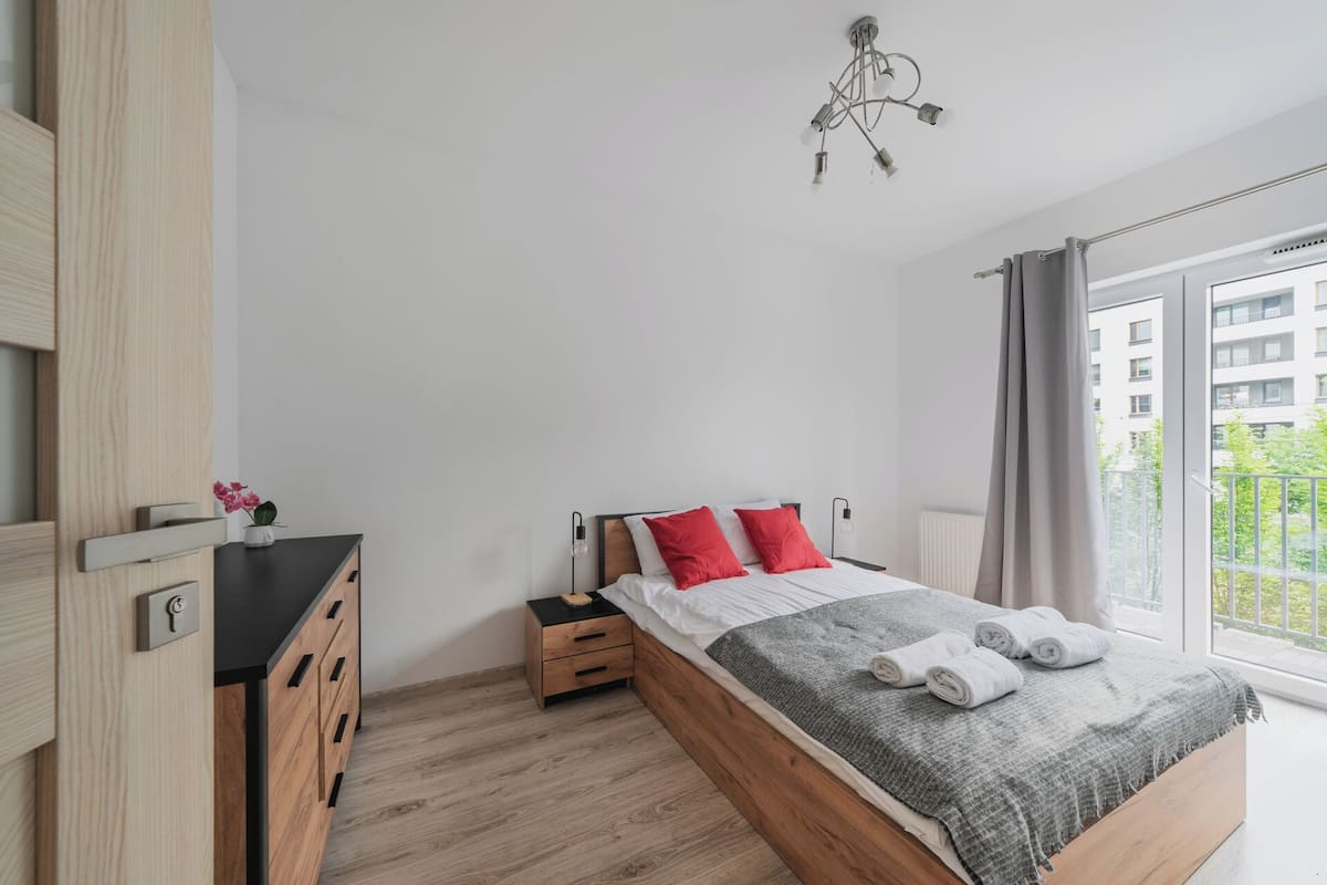 Quarto com cama de madeira, mesa de cabeceira, cômoda, lustre e vista para prédios pela janela.