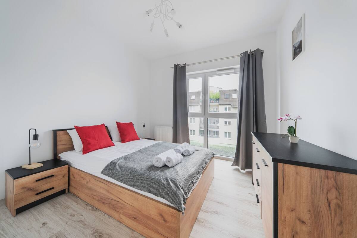 Quarto com cama de madeira, almofadas vermelhas, mesa de cabeceira com abajur, cômoda e janela com cortinas.