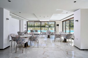 Espaço interno moderno com vista para a piscina, com cadeiras e mesas estampadas, e uma parede de vidro.