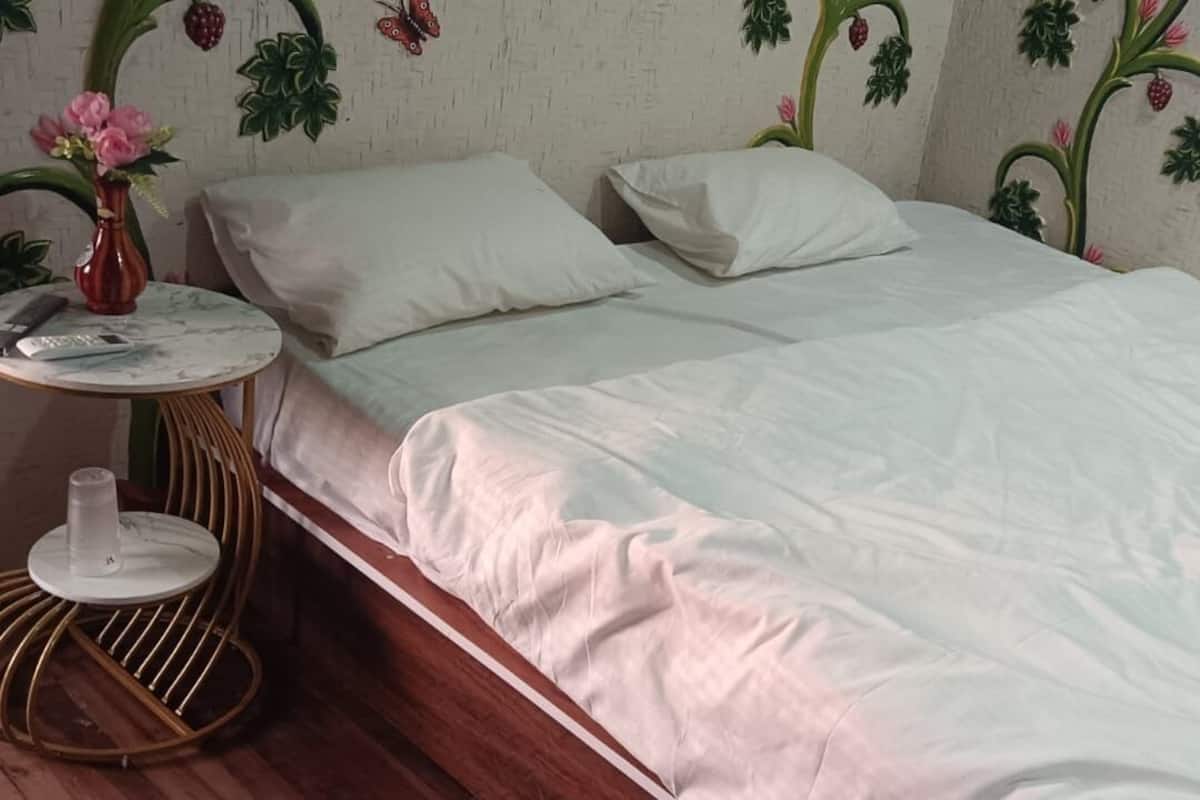 Uma cama de solteiro com roupa de cama branca, uma mesinha redonda com um vaso de flores e uma cadeira com almofada vermelha.
