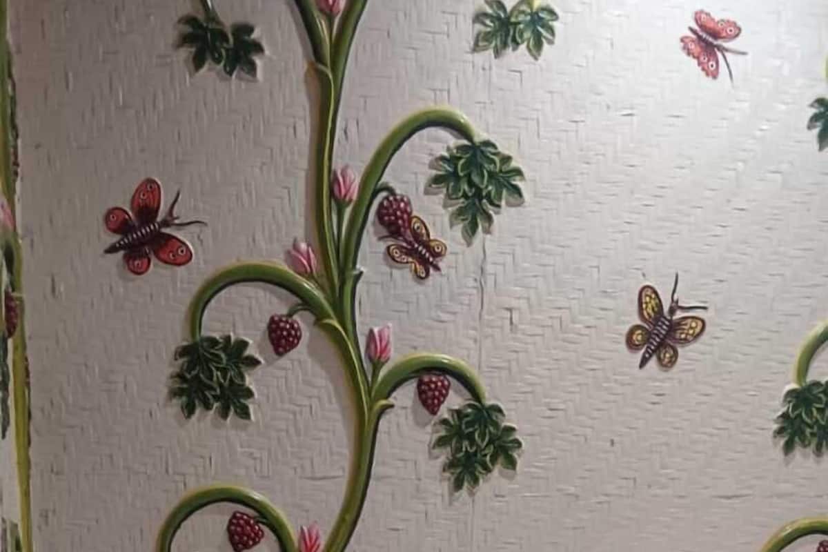 Uma cama com roupa de cama branca, um vaso vermelho com flores cor-de-rosa e uma parede com desenhos florais e de borboletas.