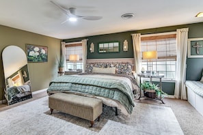 Quarto com cama, um banquinho, mesa de cabeceira com abajur, janela com cortinas e espelho com obras de arte na parede.