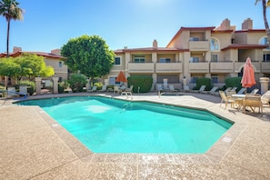 Piscina com espreguiçadeiras e guarda-sóis, rodeada por um edifício com varandas e céu ensolarado.