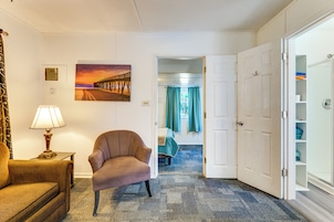 Quarto de hotel com uma cadeira, um abajur, um quadro e uma porta aberta para outro cômodo.