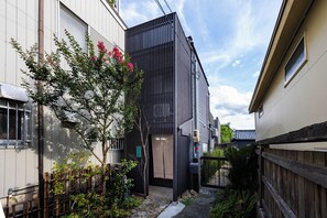 Um beco urbano estreito com um edifício moderno com ripas verticais, uma árvore florida em tons de rosa e um portão de estilo japonês tradicional.