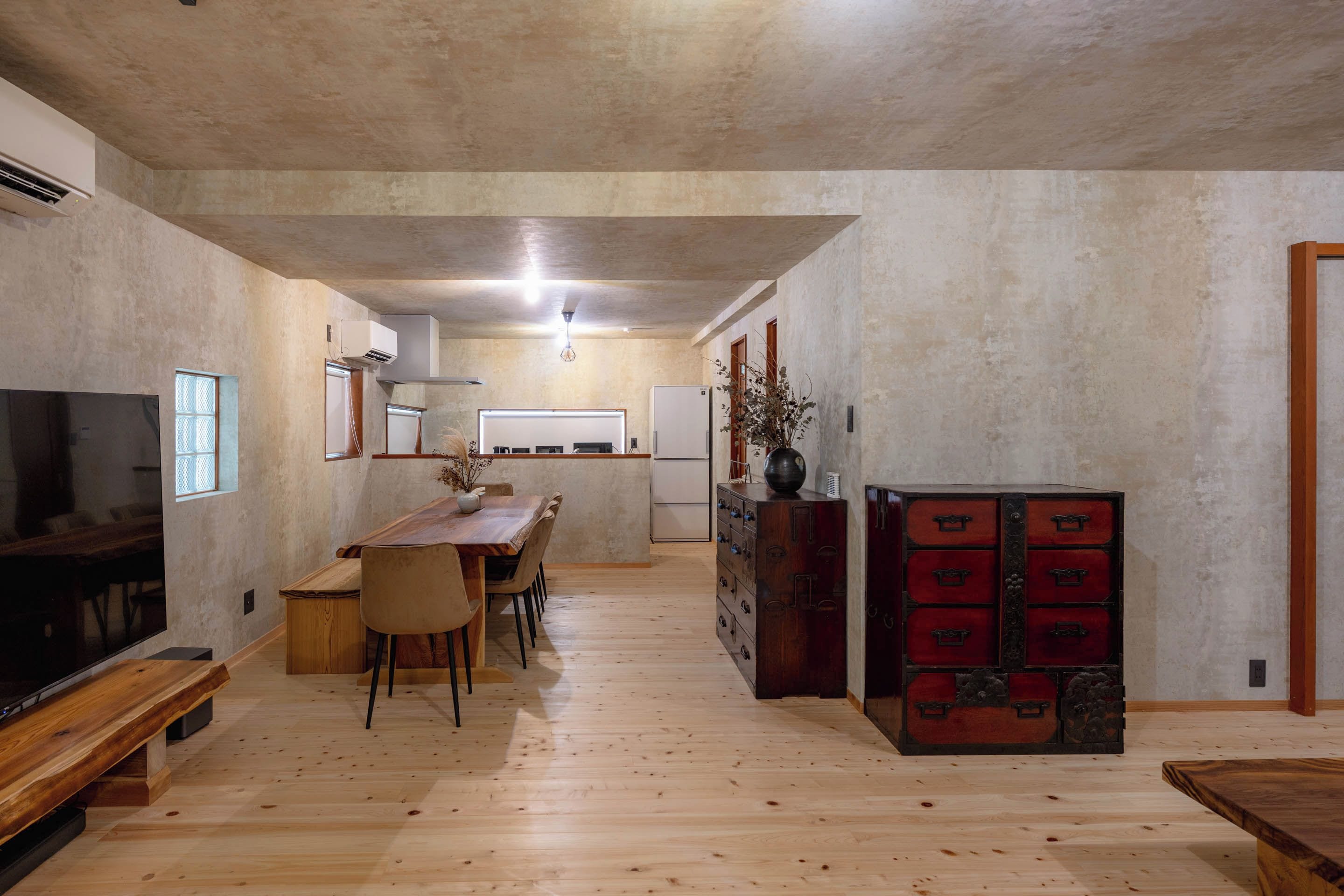 Ampla sala de estar com mesa de jantar de madeira, cadeiras, televisão e cozinha ao fundo.