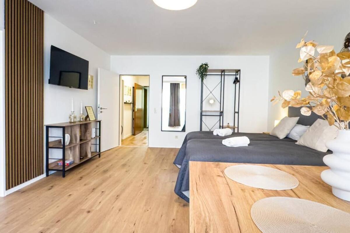 Quarto moderno com piso de madeira, cama com roupa de cama escura, televisão na parede e mesa de cabeceira de madeira com um vaso de flores secas.