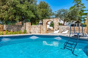 Piscina com paredes de pedra, um arco de pedra e cadeiras de descanso.
