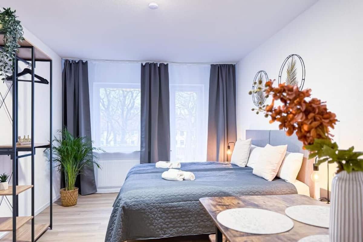 Quarto de hotel moderno com cama grande, mesa de cabeceira, pequena escrivaninha, planta em vaso e vista para o exterior.