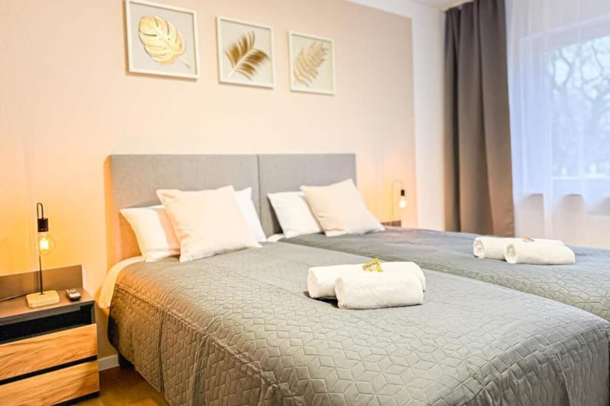 Quarto de hotel com cama, almofadas, mesa de cabeceira, luminária e arte com folhas emolduradas na parede.