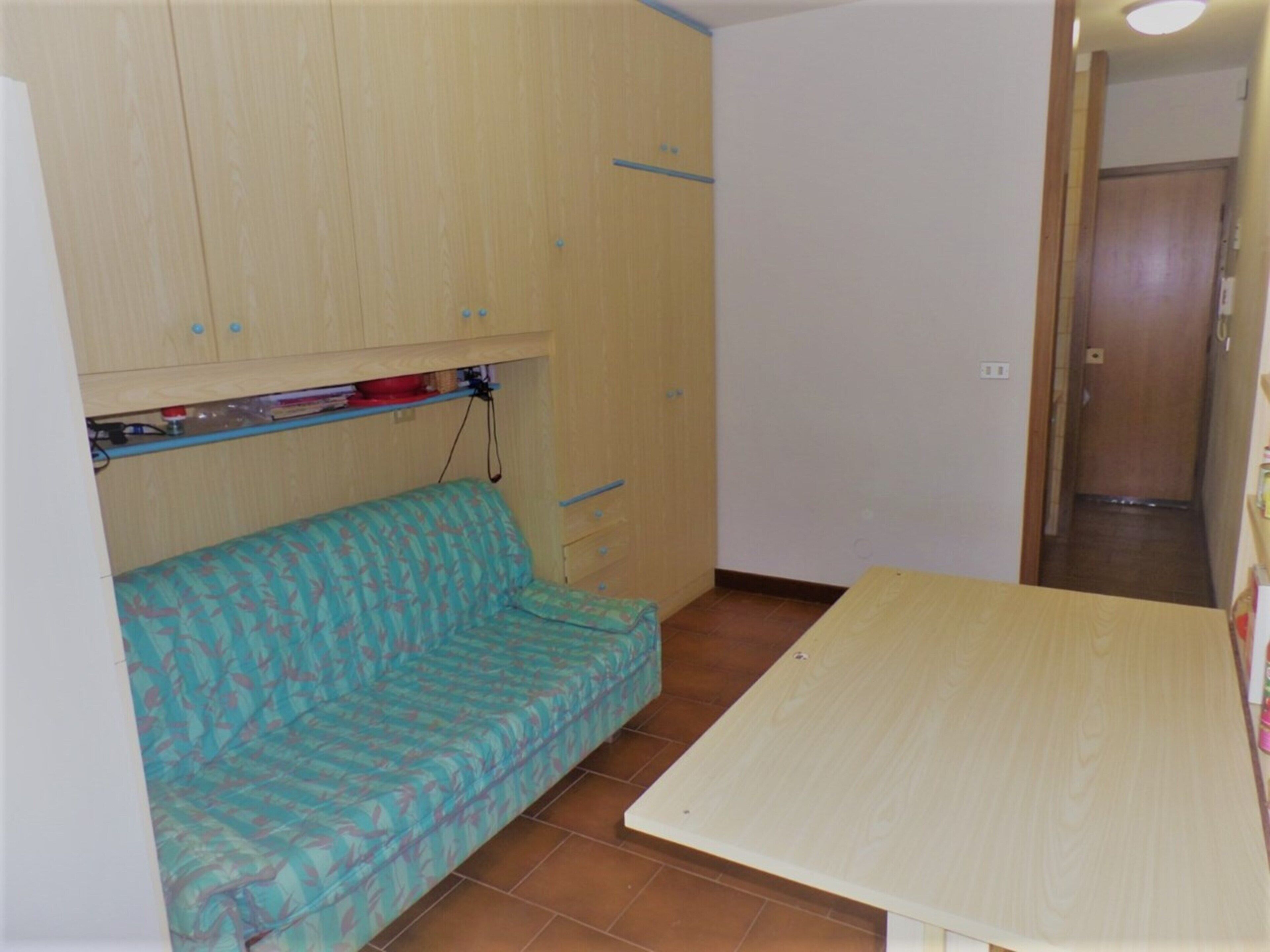 Quarto pequeno com sofá, mesa de madeira e porta para outro cômodo.