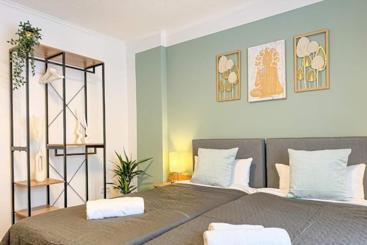 Uma cama de casal com colcha cinza, um aparador de madeira com roupas penduradas e um vaso com planta.