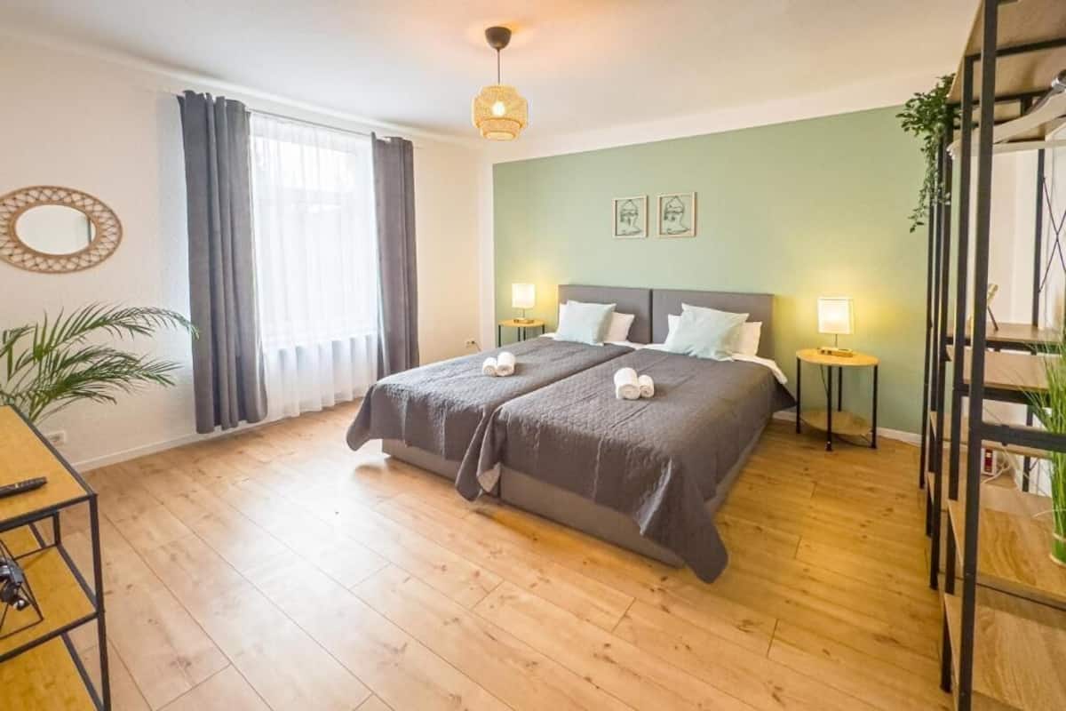 Quarto com cama de casal, mesas de cabeceira, luminária suspensa, espelho e janela com cortinas.