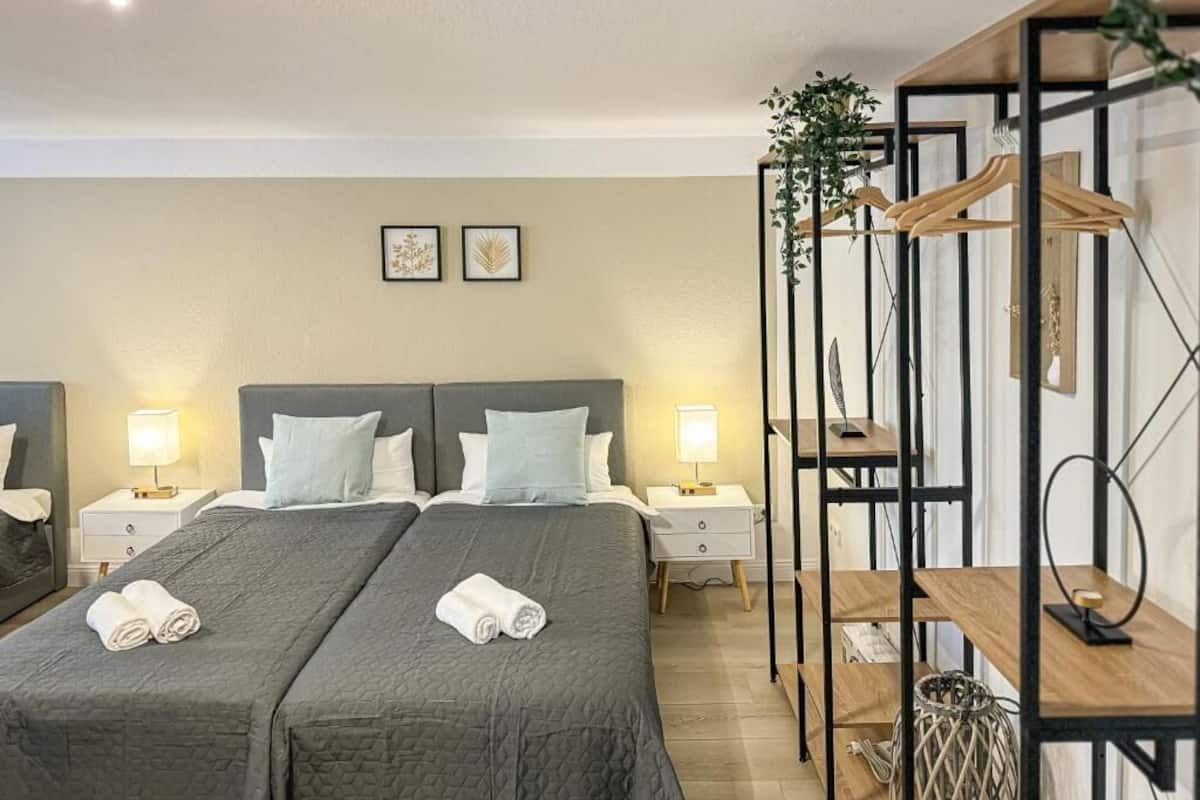 Quarto de hotel com duas camas, mesas de cabeceira com abajures, um guarda-roupa com espelho e uma planta na parede.