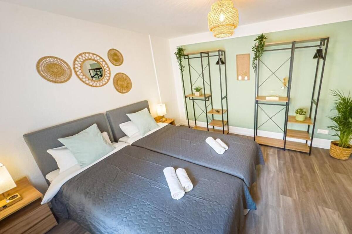 Uma cama de casal com cobertura acolchoada, duas mesas de cabeceira com abajures, uma prateleira na parede com plantas e um lustre.