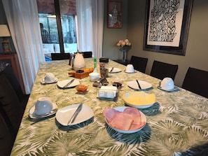 Uma mesa de jantar posta com pratos, talheres e acompanhamentos, com vista para o exterior através de amplas janelas.