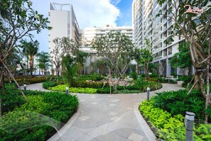 Um pátio urbano moderno com vegetação, um espelho d'água central e edifícios contemporâneos ao fundo.
