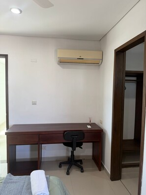 Quarto com escrivaninha de madeira, uma cadeira, cama com toalha enrolada, ar condicionado e guarda-roupa.