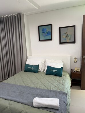 Cama bem arrumada com duas almofadas, um cobertor e uma toalha dobrada. Há duas obras de arte emolduradas na parede acima da cama.