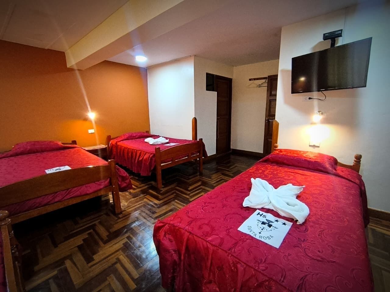 Quarto de hotel com três camas de solteiro, televisão presa à parede e chão de madeira.