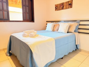 Cama bem arrumada com colcha azul e almofadas brancas, uma toalha enrolada sobre a cama e uma janela com persianas de madeira.
