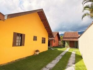 Um edifício amarelo com telhado inclinado, uma pequena área de jardim com um caminho e uma montanha ao fundo.