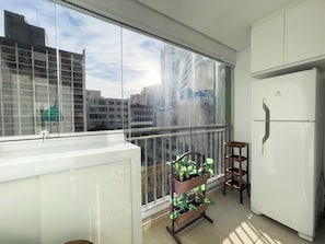 Uma varanda com vista para edifícios, um refrigerador branco e um suporte para planta.