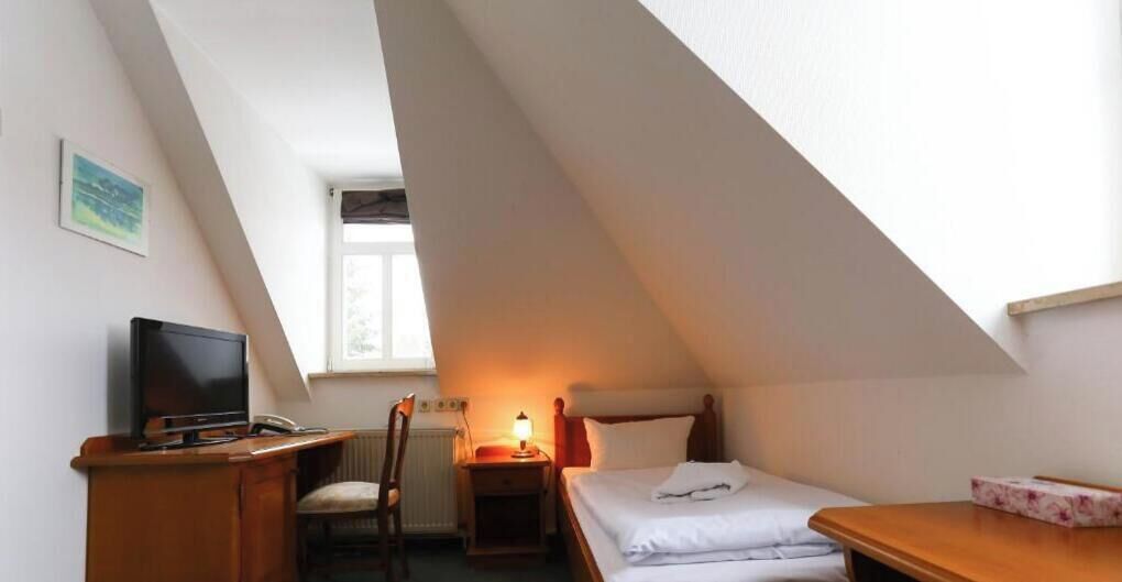 Um quarto pequeno e triangular com uma cama, uma escrivaninha, uma cadeira, uma televisão e uma janela.