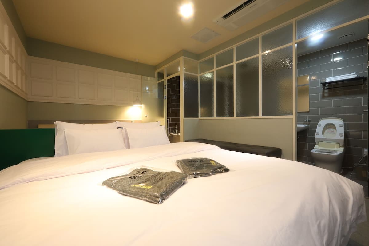 Quarto de hotel com cama, banheiro com vaso sanitário e pia, e janela com vidro fosco.