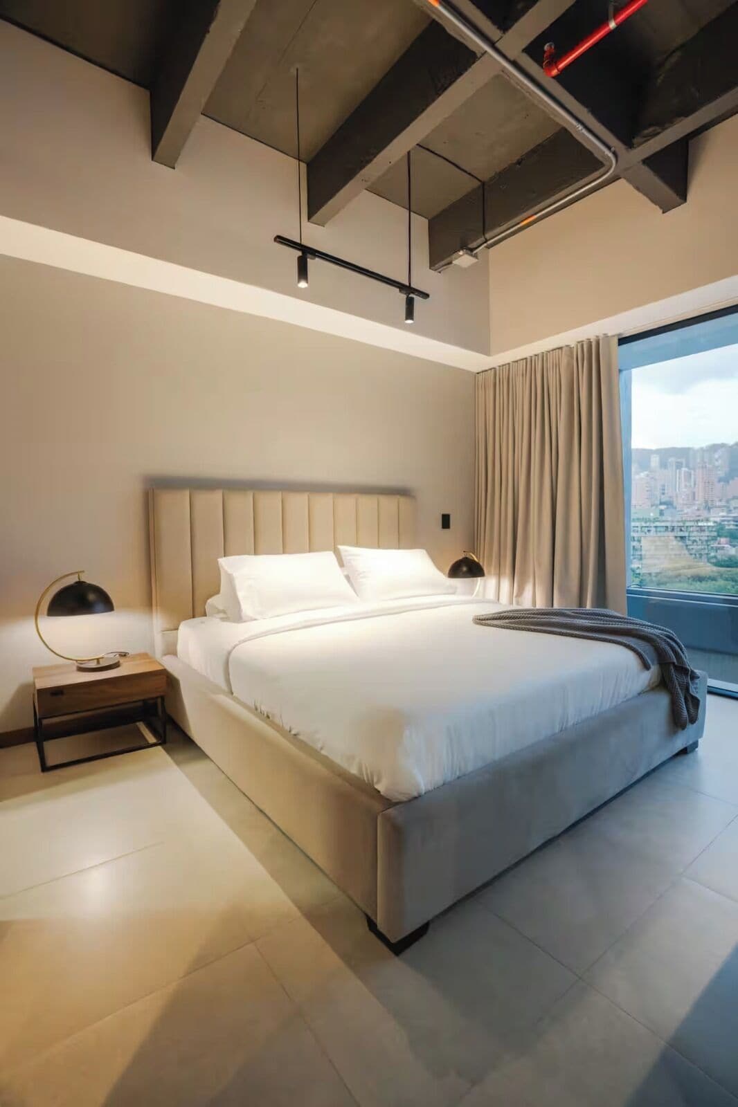 Quarto moderno com uma cama grande, uma mesinha de cabeceira, uma luminária e vista para a cidade através de uma janela ampla.