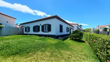 Uma casa de dois andares com fachada branca e persianas escuras, rodeada por vegetação bem cuidada.