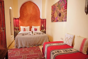 Quarto de hotel com uma grande porta arqueada, uma cama com colcha de padrão vermelho e branco, uma cama menor com manta vermelha e branca, e uma tapeçaria colorida na parede.
