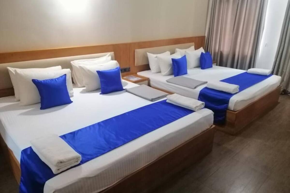 Quarto de hotel com duas camas, cada uma com roupa de cama azul e almofadas brancas, uma cabeceira de madeira e uma mesa de cabeceira com abajur.