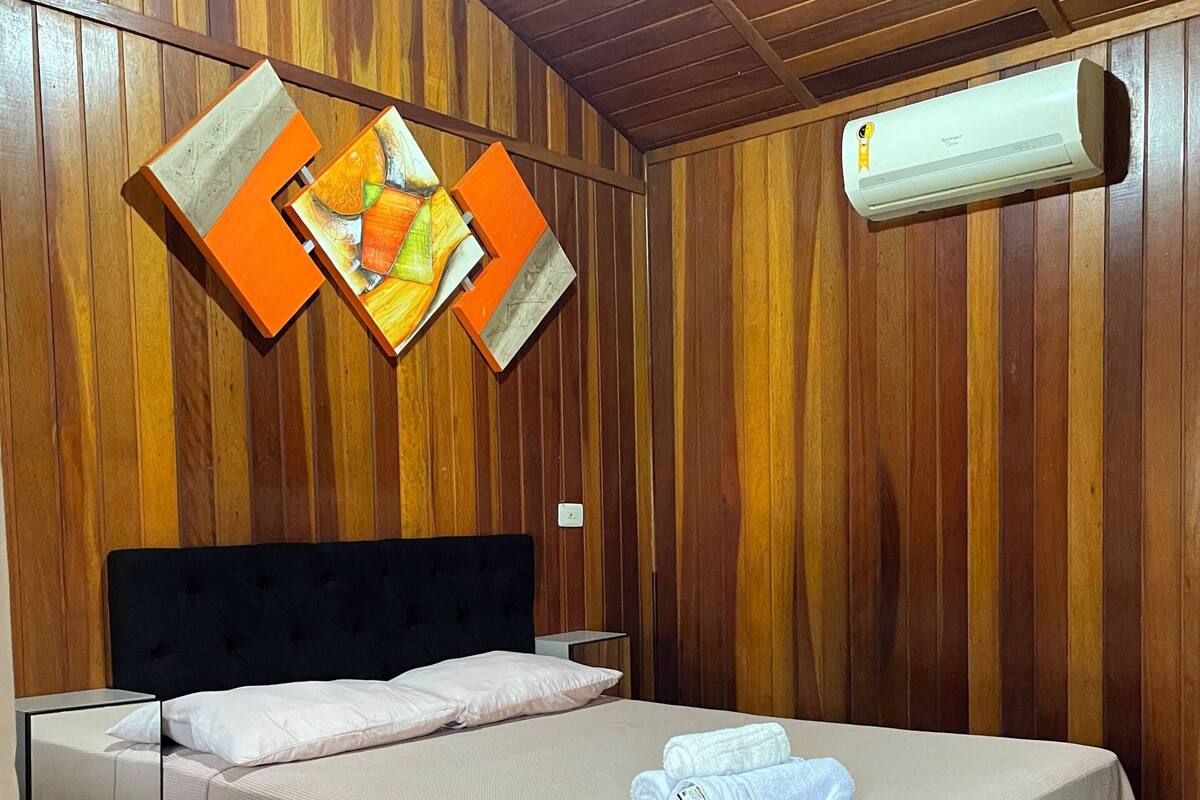 Quarto de hotel com parede revestida em madeira, cama com cabeceira preta, almofadas brancas e roupa de cama bem arrumada com toalhas.