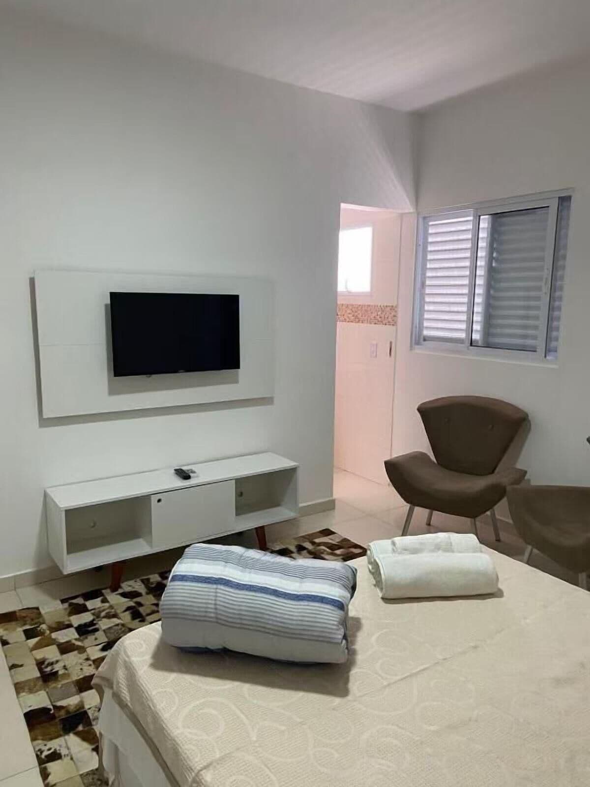 Quarto com cama, televisão na parede, uma cadeira e janela com persianas.