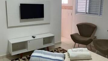 Quarto com cama, televisão na parede, uma cadeira e janela com persianas.