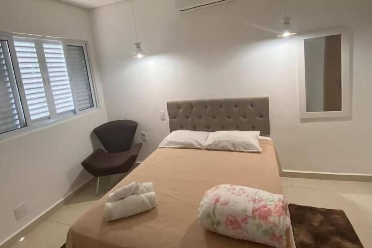 Um quarto com cama, uma cadeira, janela com persianas, ar condicionado e ventilador de teto.