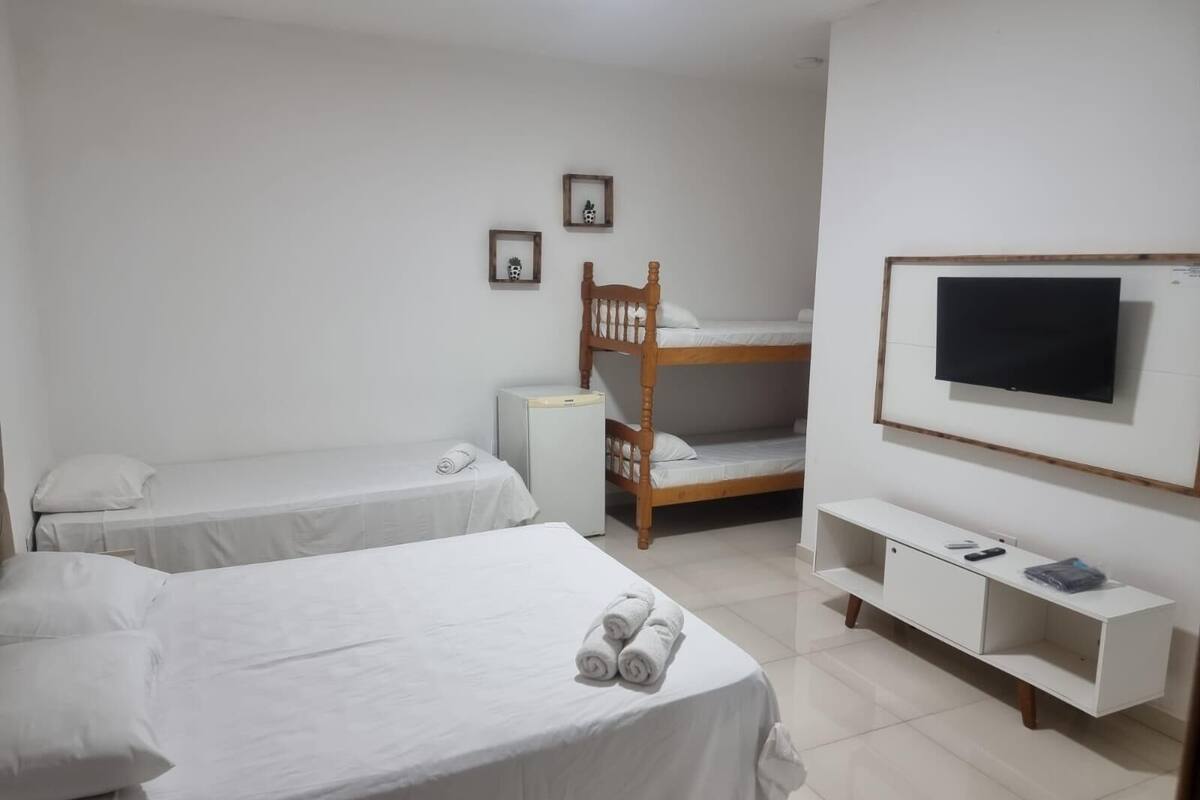 Quarto com duas camas de solteiro, uma cama beliche, televisão, pequena escrivaninha e prateleira.