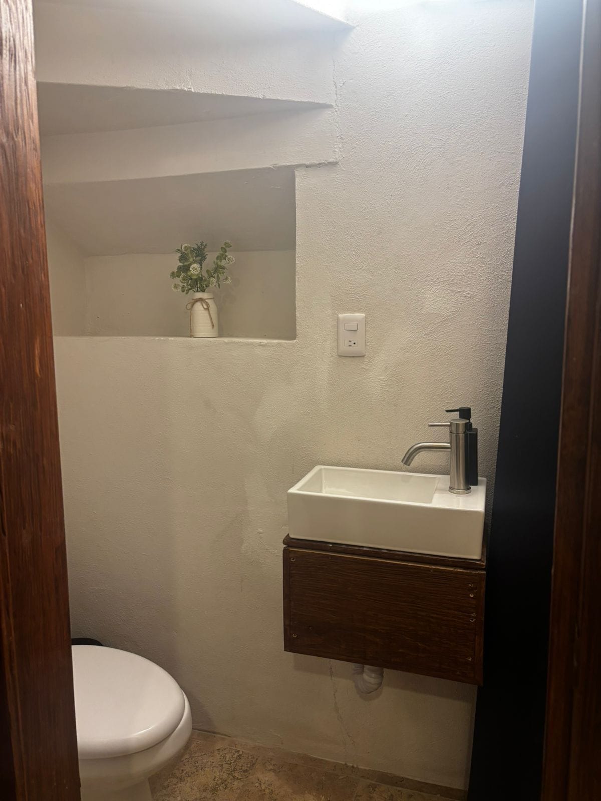 Um banheiro pequeno com vaso sanitário, pia com torneira, espelho e prateleira com uma planta.