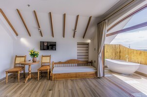 Quarto com cama, duas cadeiras, mesa com um vaso de flores, televisão e uma grande janela com vista.
