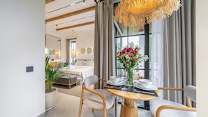 Quarto de hotel moderno com cama, área de refeições com mesa e cadeiras, janela grande com cortinas e luminária pendente.