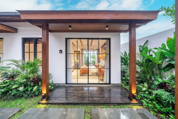 Uma casa moderna com entrada coberta, um quarto visível com cama e cortinas, e vegetação exuberante ao redor.