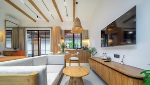 Uma sala de estar moderna com sofá, mesa redonda de madeira, televisão na parede e área de cozinha com armários de madeira e uma janela.