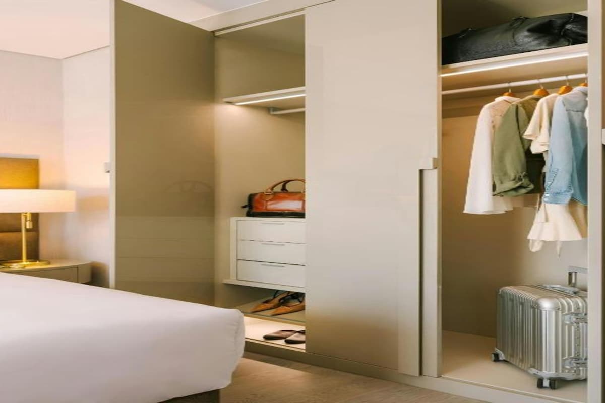 Quarto de hotel com cama, luminária de cabeceira, guarda-roupa com roupas penduradas, mala e radiador.