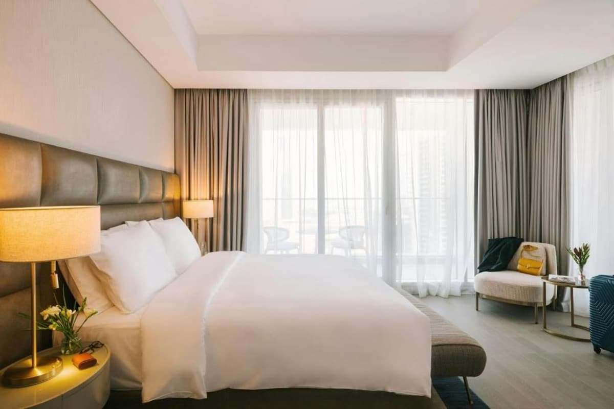 Quarto de hotel com uma cama grande, abajures, uma cadeira, uma mesinha e vista para o exterior através de cortinas leves.